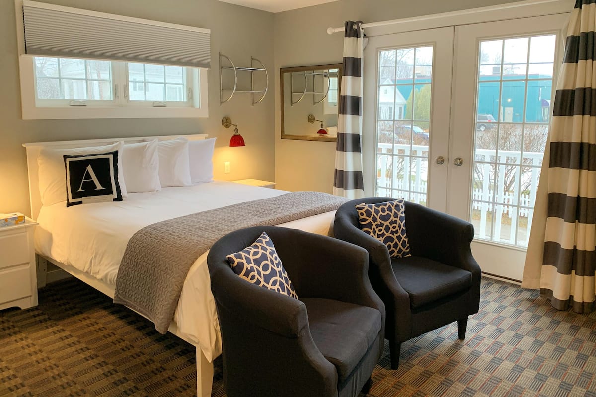 Quarto de hotel com cama, duas poltronas, mesa de cabeceira, luminária, janela com persianas, porta com vista e espelho.