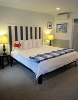 Cama bem arrumada com cabeceira listrada, duas luminárias de cabeceira, um travesseiro em formato de caranguejo vermelho e um quadro na parede.