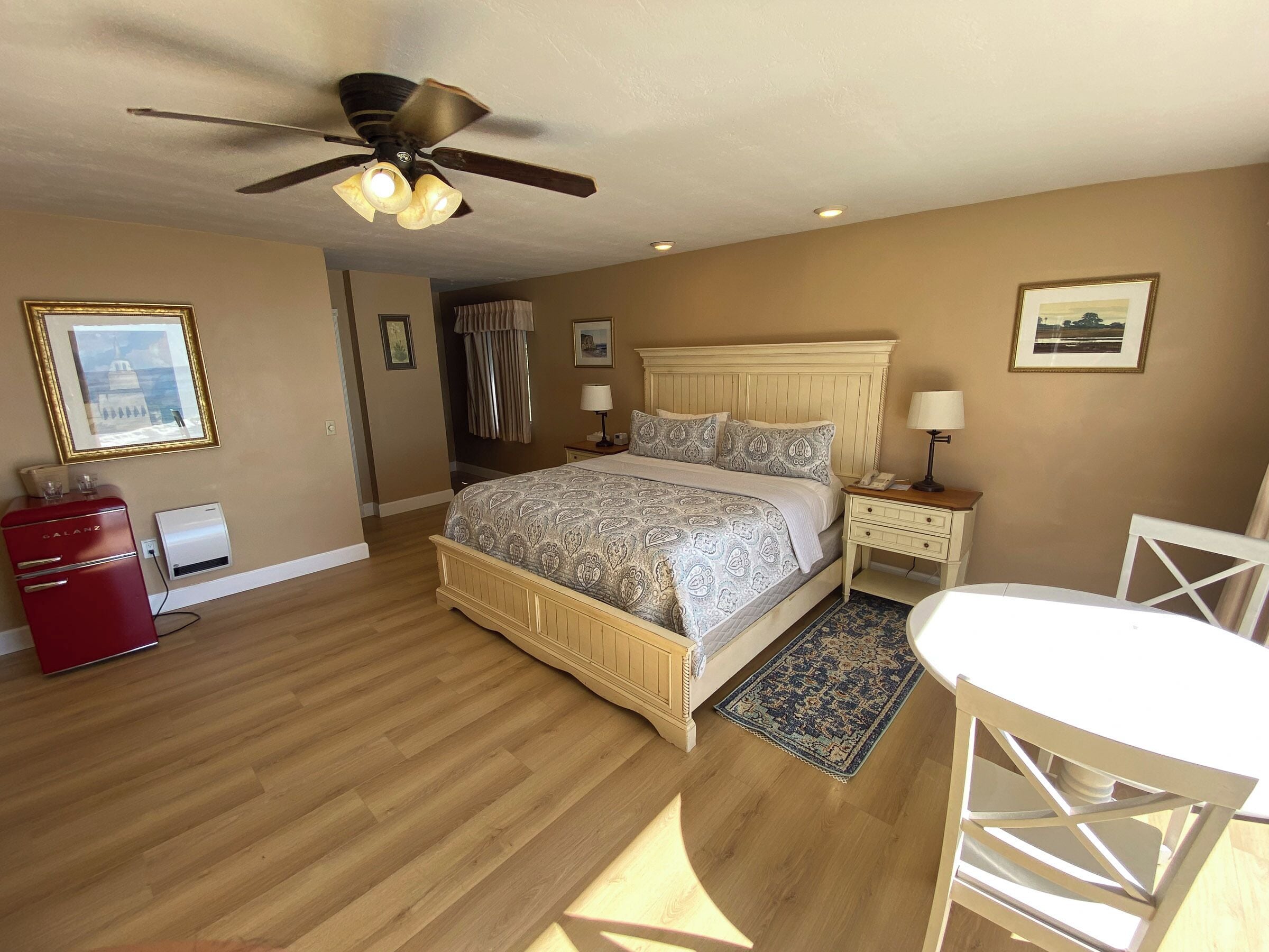 Quarto com piso de madeira, cama com roupa de cama estampada, ventilador de teto, luminárias de cabeceira, mesa de cabeceira, eletrodoméstico vermelho e um quadro na parede.