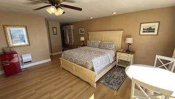 Quarto com piso de madeira, cama com roupa de cama estampada, ventilador de teto, luminárias de cabeceira, mesa de cabeceira, eletrodoméstico vermelho e um quadro na parede.