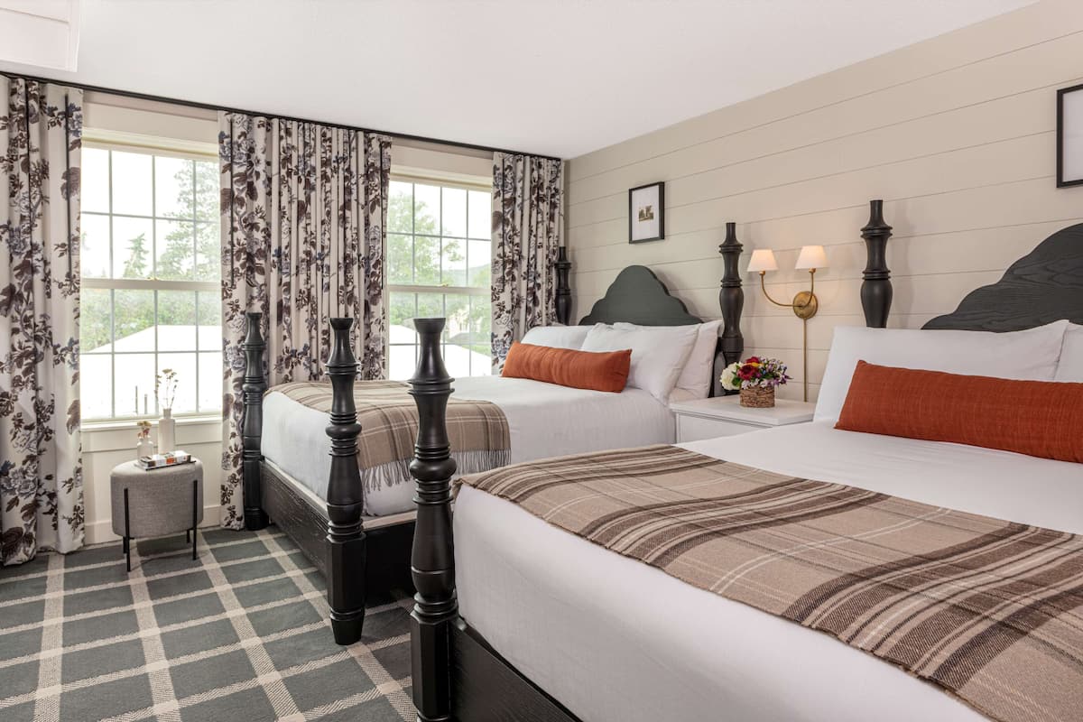 Quarto de hotel com duas camas, chão xadrez, cortinas florais e janela com vista.
