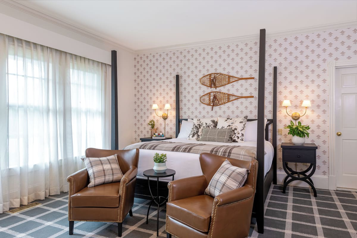 Quarto com cama de dossel, duas poltronas de couro marrom, uma mesinha redonda com uma planta e uma mesa de cabeceira com abajures.