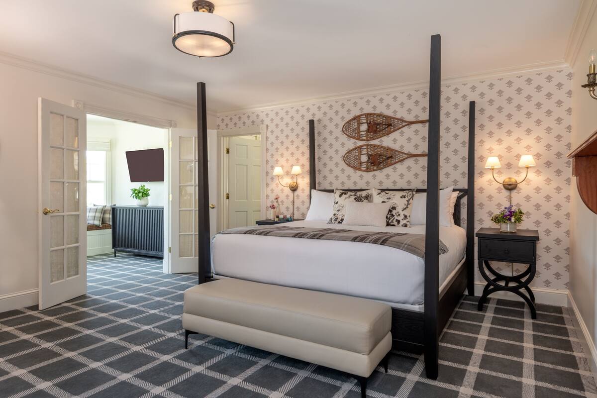 Quarto com cama de dossel, um banco, mesa de cabeceira com abajures e papel de parede estampado.