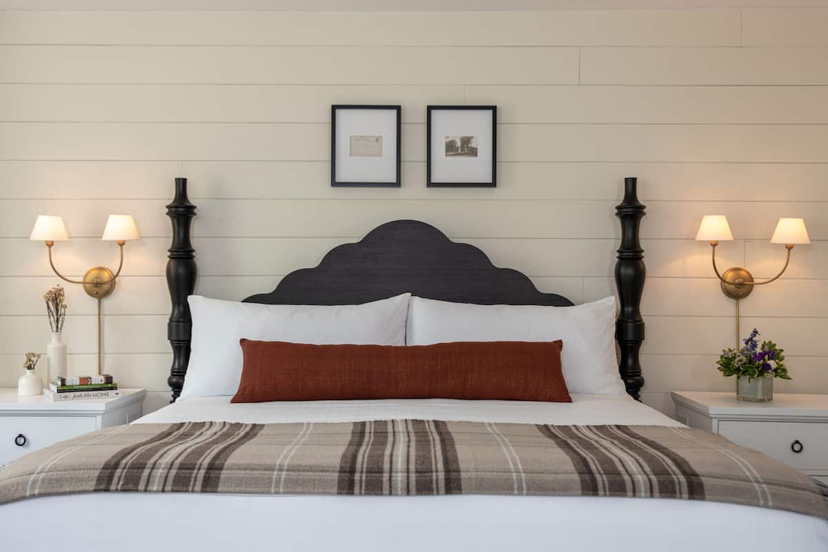 Uma cama bem arrumada com cabeceira escura, duas quadros emoldurados na parede e abajures de cada lado.
