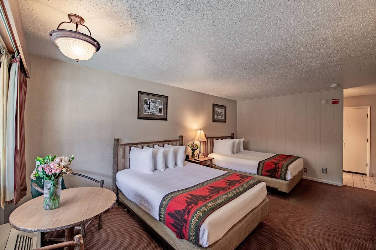 Quarto de hotel com duas camas, uma mesinha com um vaso de flores, uma luminária e quadros nas paredes.