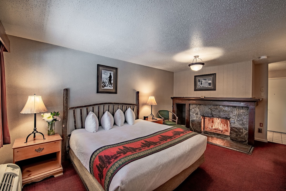 Quarto de hotel com uma cama grande, lareira de pedra e duas luminárias fixadas na parede.