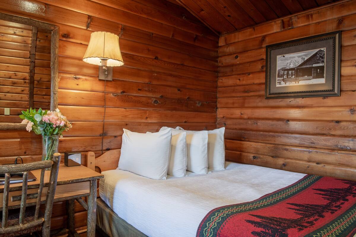 Quarto de cabana de madeira com uma cama, almofadas, uma escrivaninha com cadeira, uma luminária, um quadro na parede e um vaso com flores.