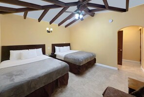 Quarto de hotel com duas camas, um ventilador de teto, uma mesa de cabeceira e uma porta que dá para outro cômodo.