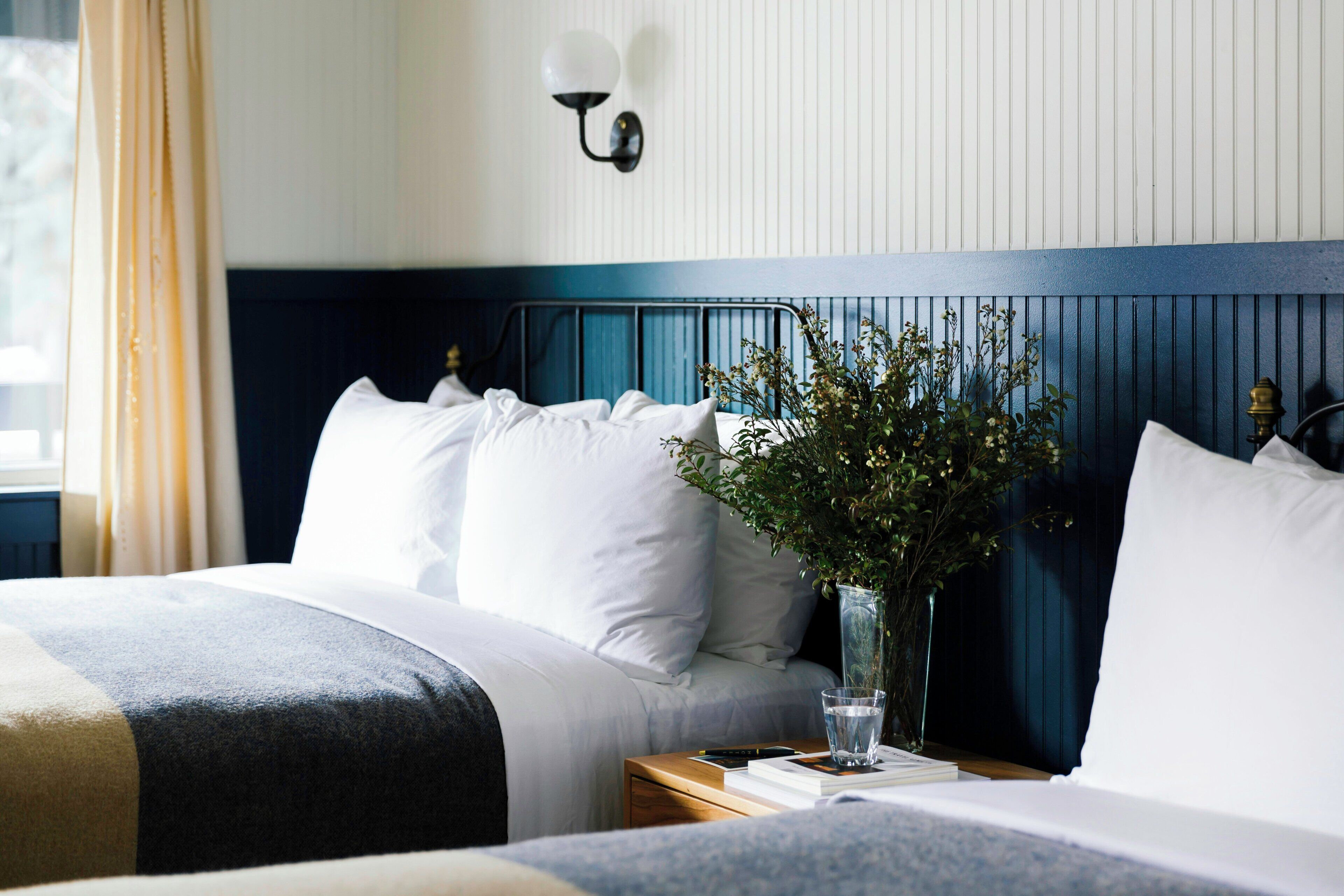 Quarto de hotel com cama, mesa de cabeceira com um copo de água, um livro e um vaso com flores.