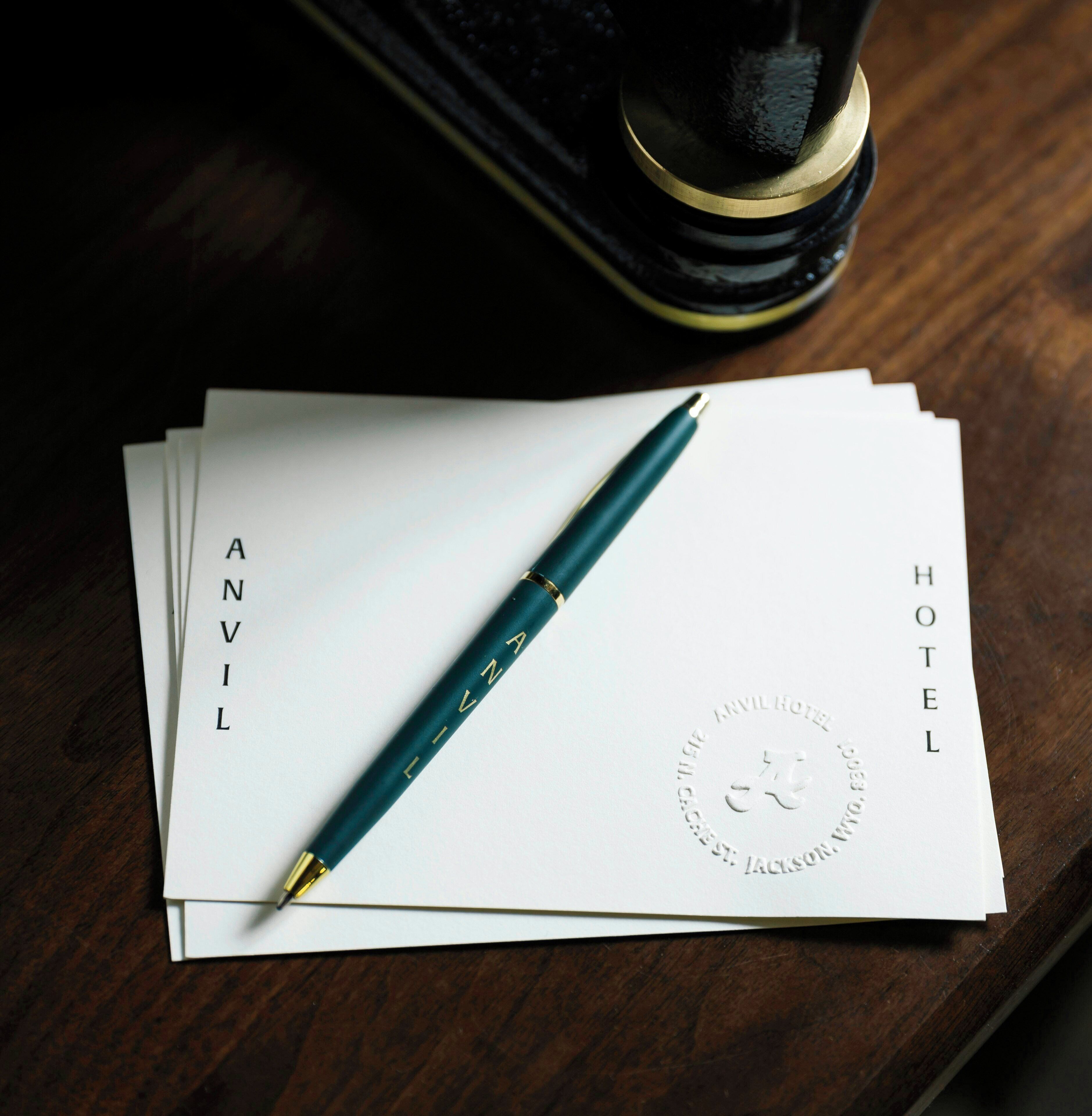Uma pilha de papéis brancos com 'ANVIL HOTEL' e 'HOTEL' impressos, uma caneta verde e uma caneta tinteiro preta sobre uma superfície de madeira.