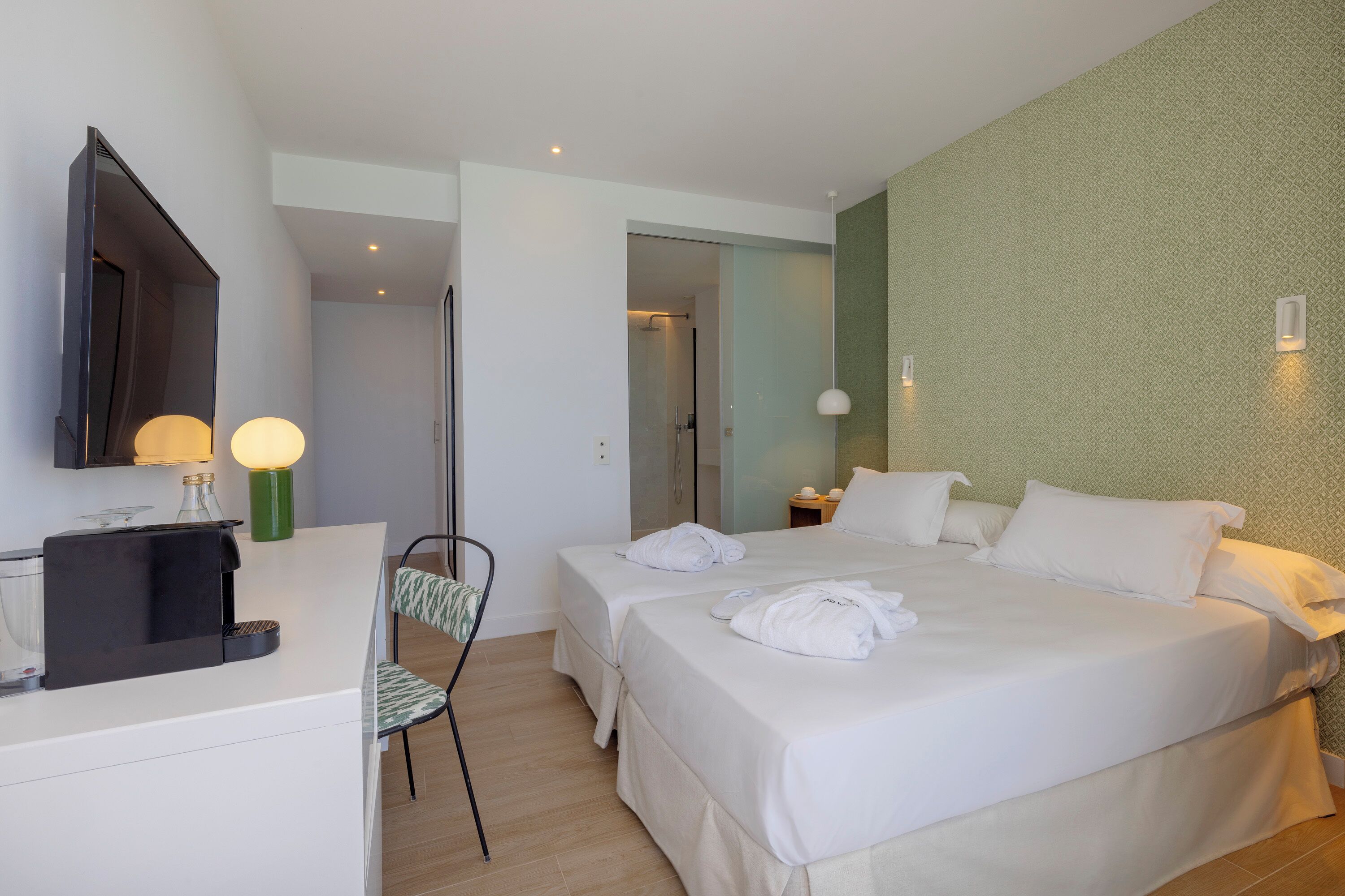 Quarto de hotel com cama, escrivaninha com cafeteira, cadeira e espelho.