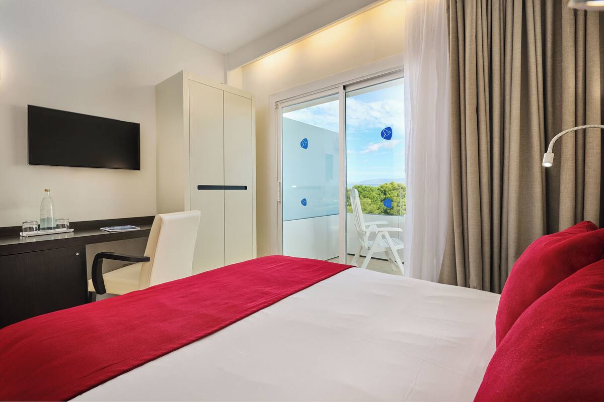 Quarto de hotel com cama, escrivaninha, cadeira, televisão e varanda com cortinas.