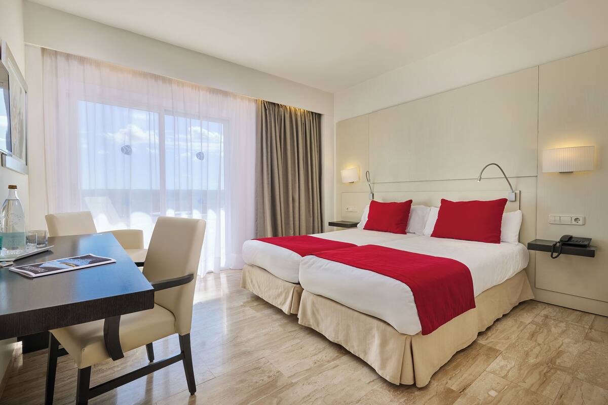 Quarto de hotel com uma cama grande, uma escrivaninha com cadeira, mesa de cabeceira com abajur e janela com cortinas.