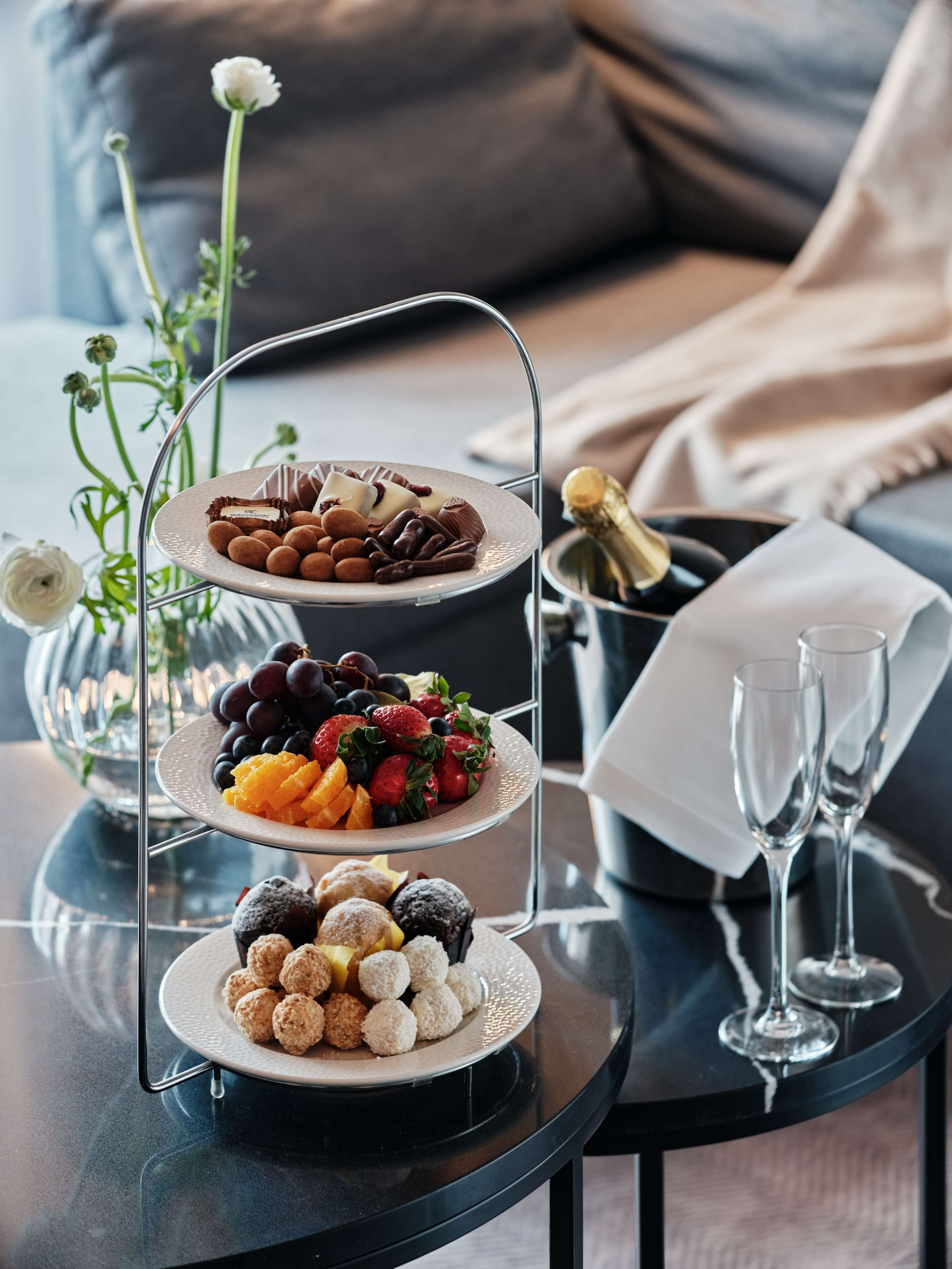 Suporte de sobremesas em três níveis com doces variados e frutas, uma garrafa de champanhe e duas taças sobre uma mesa.