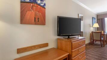 Quarto de hotel com televisão sobre uma cômoda de madeira, uma escrivaninha com cadeira e um quadro de um edifício na parede.