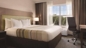 Quarto de hotel com uma cama grande, uma escrivaninha, uma cadeira, uma janela com cortinas e vista para o verde lá fora.