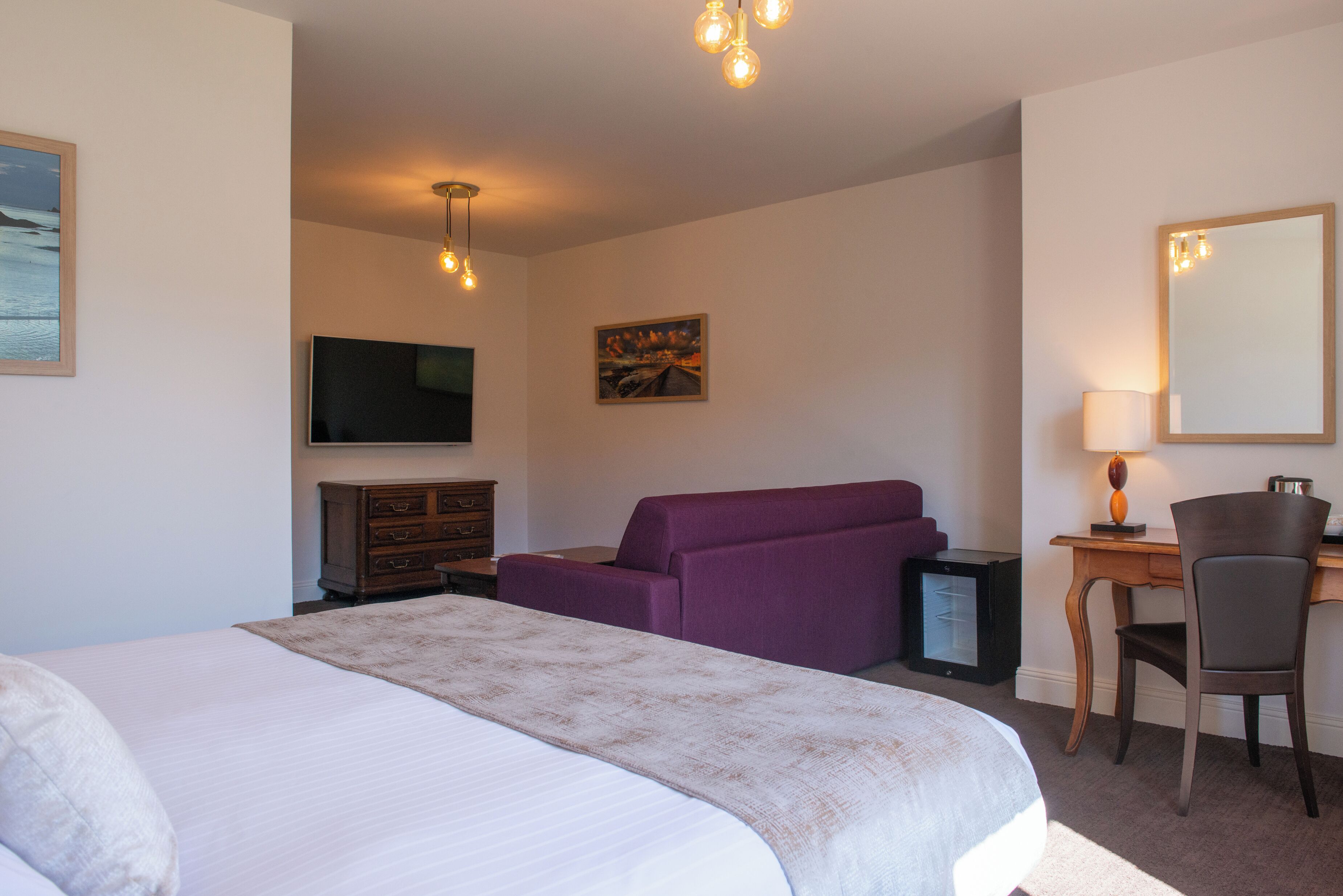 Quarto de hotel com uma cama grande, uma escrivaninha com cadeira, um sofá roxo, uma televisão e um quadro na parede.