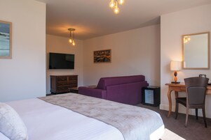 Quarto de hotel com uma cama grande, uma escrivaninha com cadeira, um sofá roxo, uma televisão e um quadro na parede.