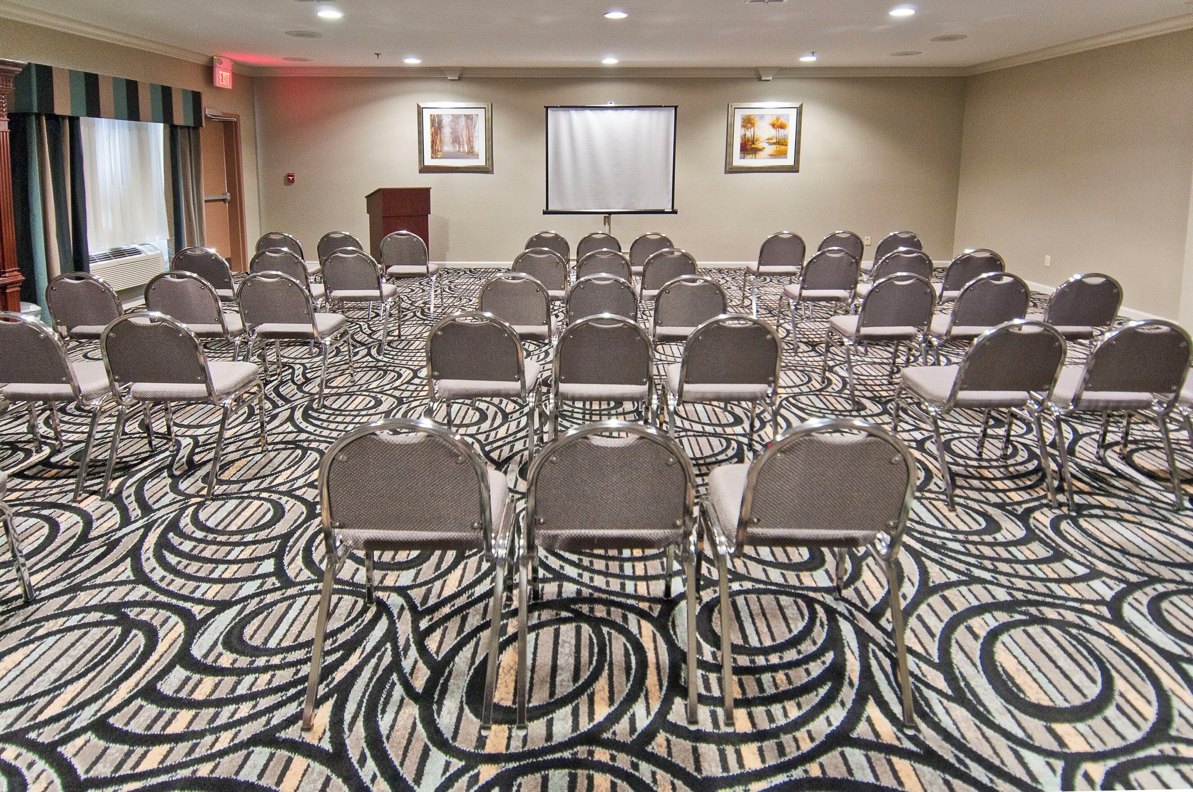 Sala de conferência com fileiras de cadeiras, uma tela de projetor e duas fotografias emolduradas na parede.