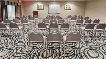 Sala de conferência com fileiras de cadeiras, uma tela de projetor e duas fotografias emolduradas na parede.