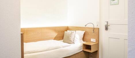 Quarto pequeno e bem organizado, com uma cama de solteiro, cabeceira de madeira, mesa de cabeceira e um tapete verde estampado.