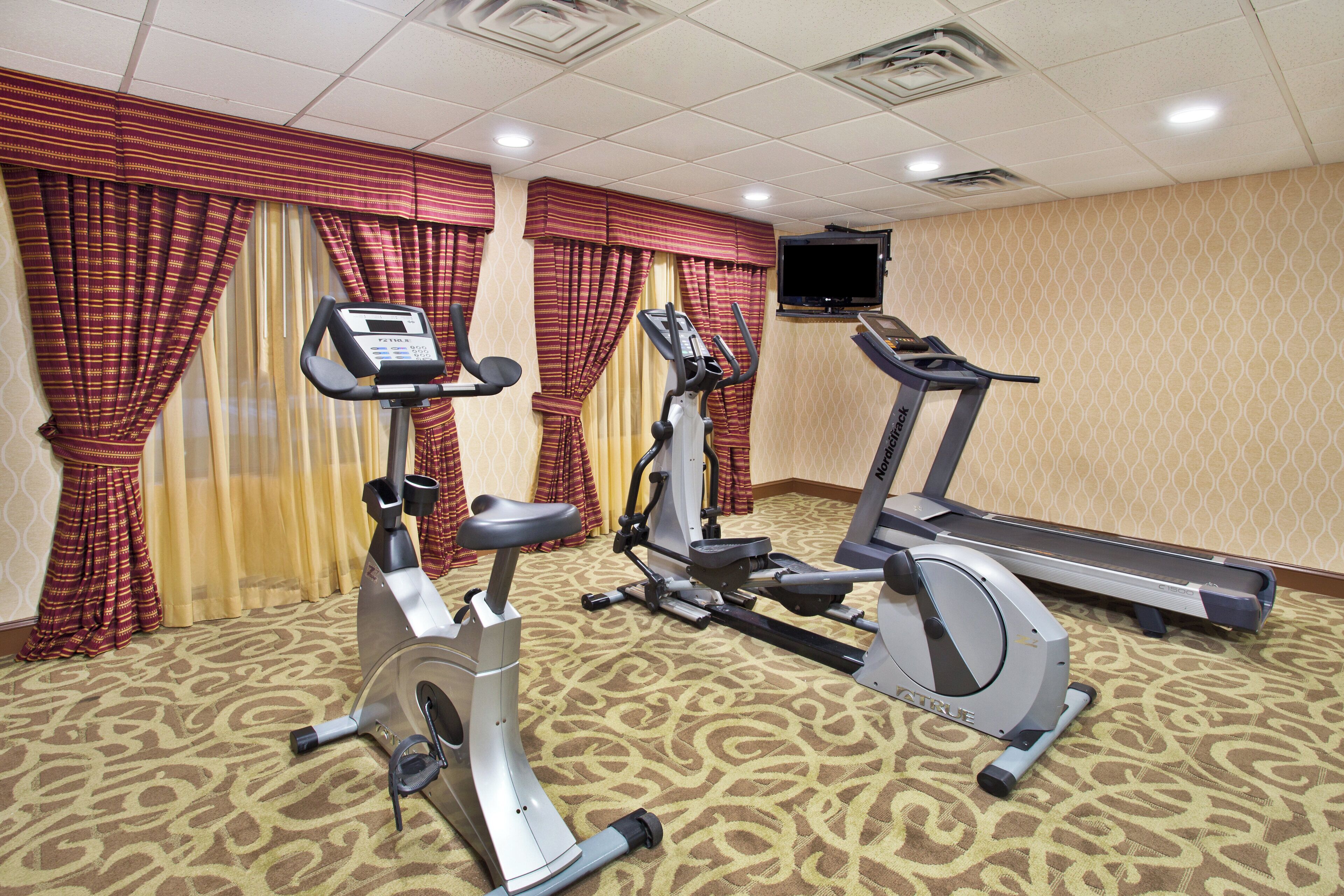 Ginásio de hotel com máquinas de cardio, televisão e carpete estampado.