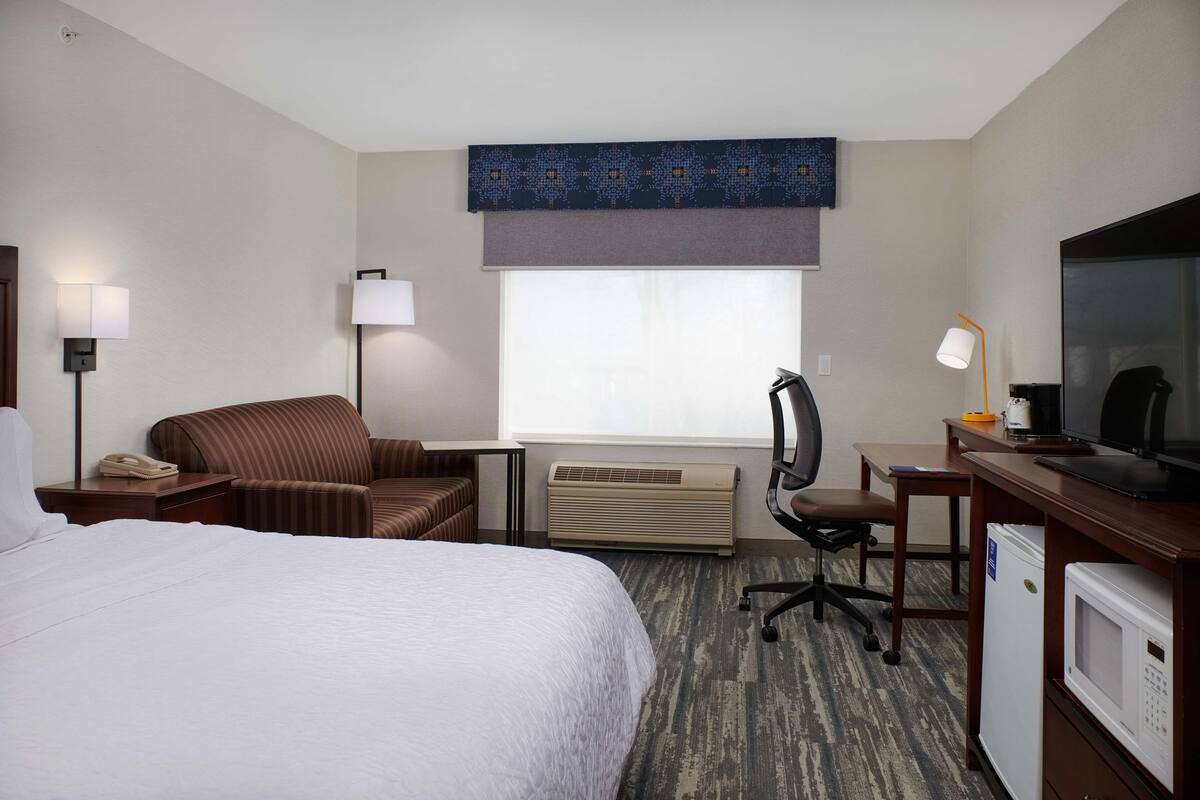 Quarto de hotel com cama, escrivaninha com computador, cadeira, televisão e micro-ondas.