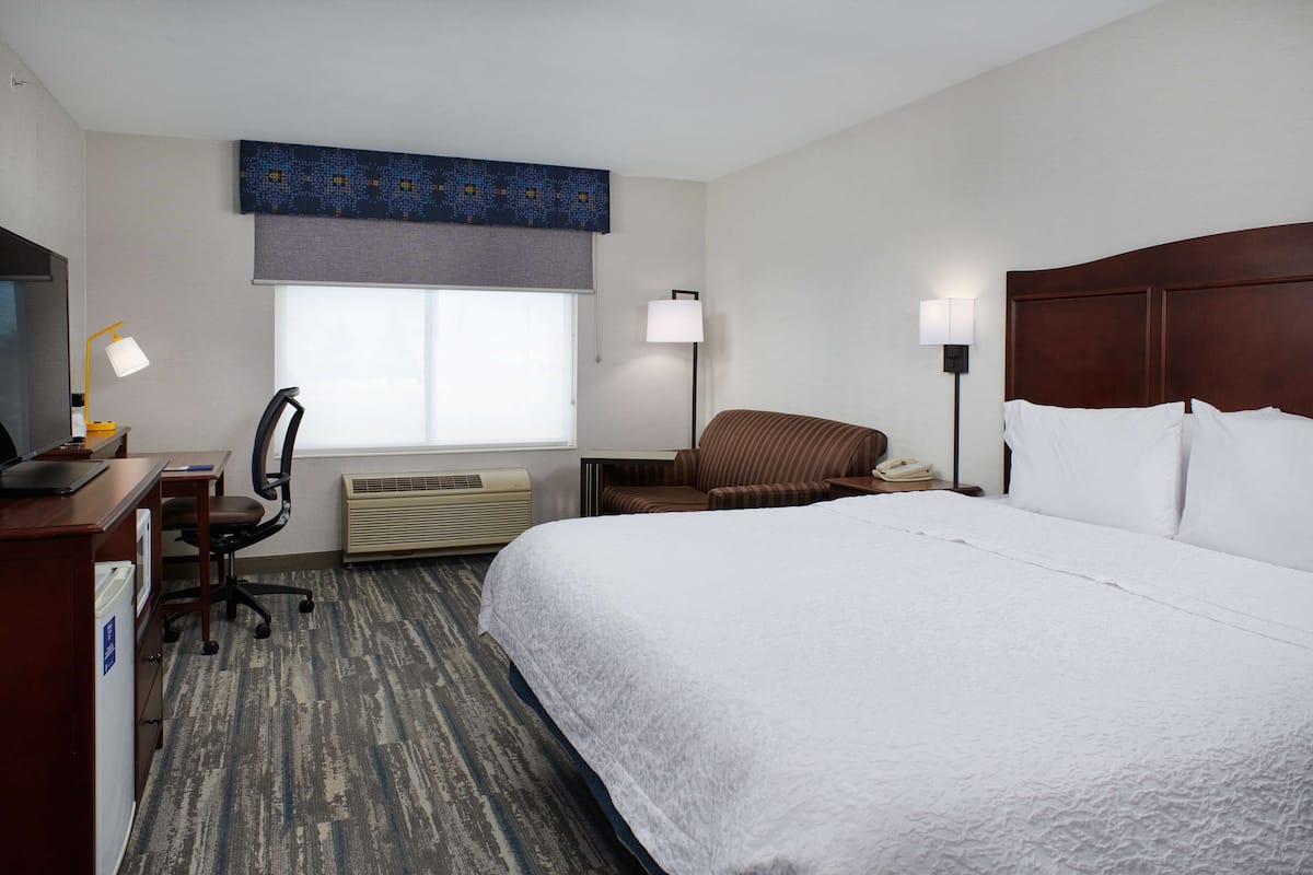 Quarto de hotel com uma cama grande, uma escrivaninha com computador, uma cadeira, uma janela com persianas e um abajur fixado na parede.