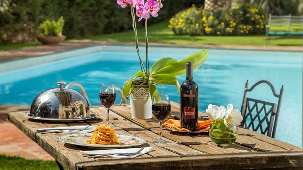 Uma mesa de jantar com pratos de comida, copos de vinho, uma garrafa de vinho e um vaso de flores junto a uma piscina.