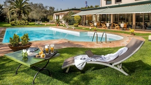 Uma área na piscina com espreguiçadeira, mesa com bebidas e um edifício com assentos externos.