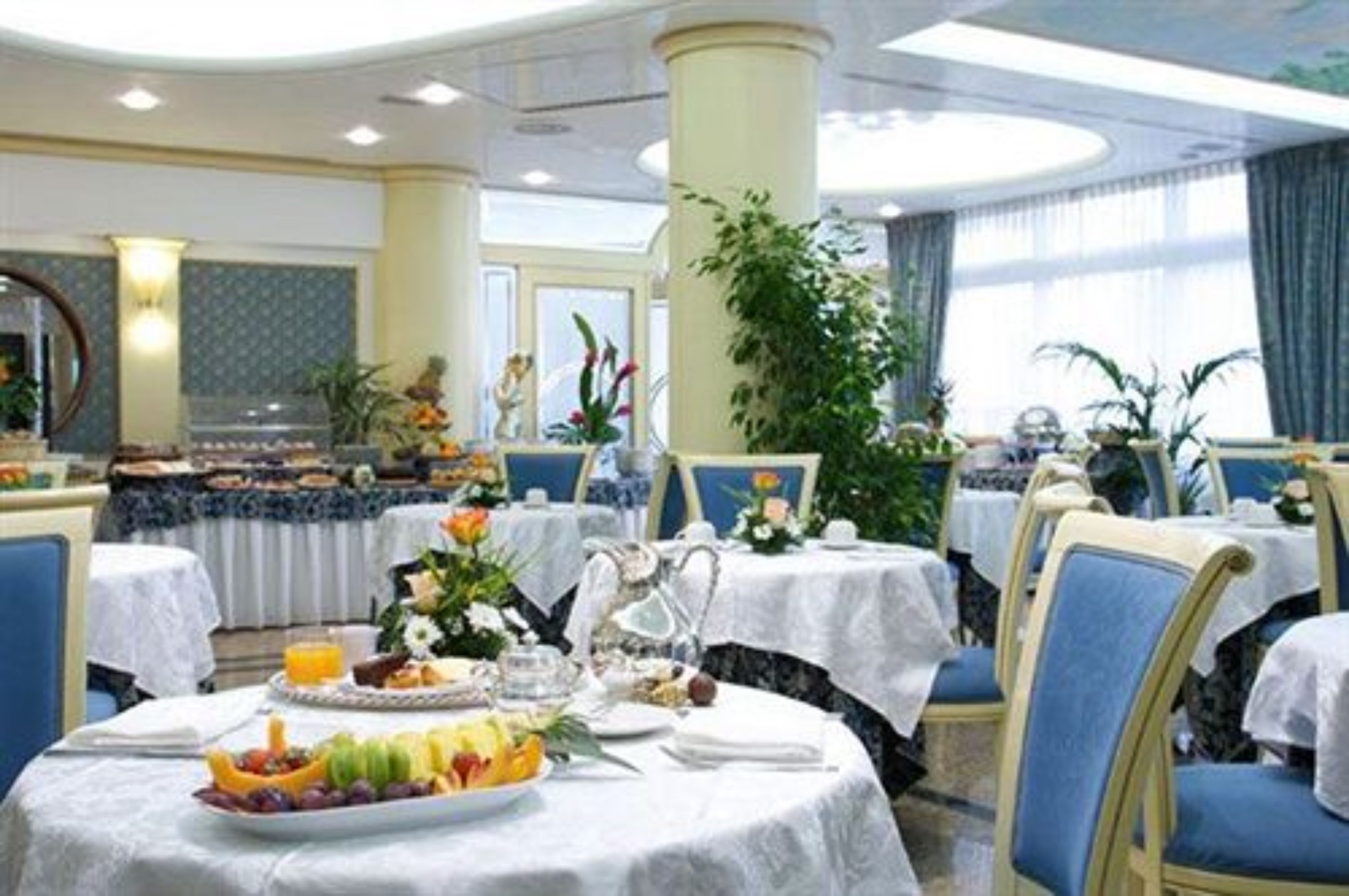 Uma sala de jantar de hotel com mesas redondas, cadeiras azuis e toalhas de mesa brancas.