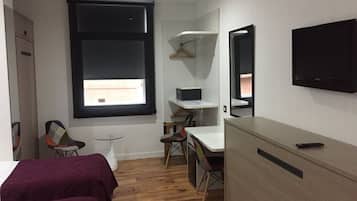 Quarto de hotel pequeno com uma cama, uma escrivaninha, uma cadeira, uma televisão e uma janela.