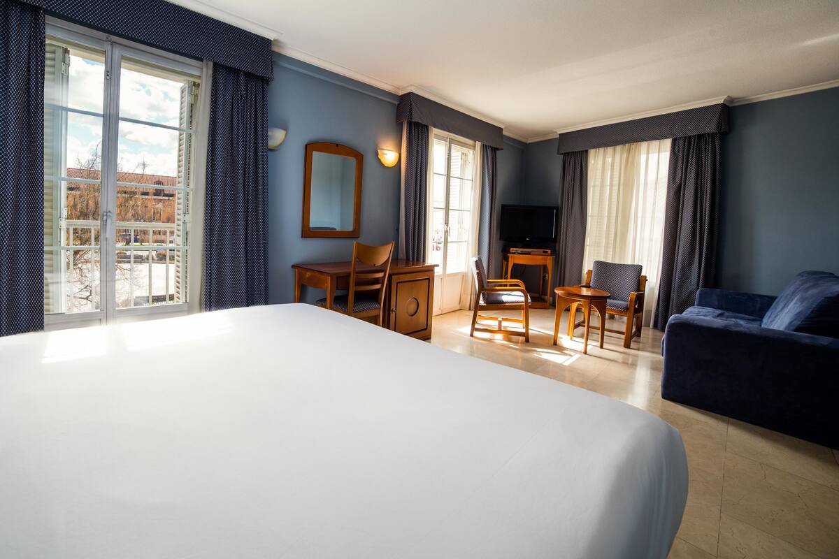Quarto de hotel com uma cama grande, uma escrivaninha com cadeira, um sofá e uma televisão.