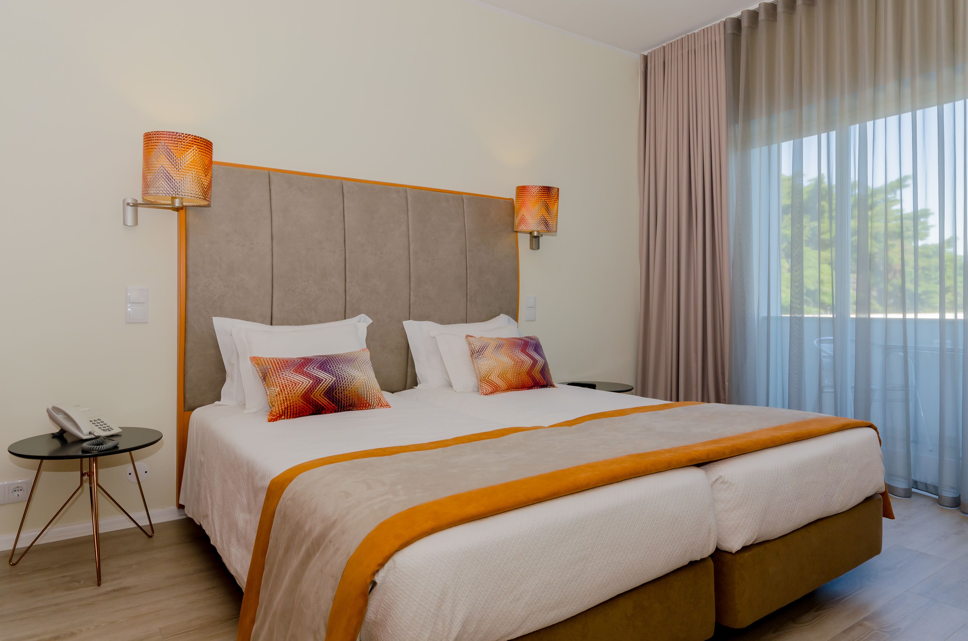 Quarto de hotel com uma cama grande, duas luminárias de cabeceira, uma mesinha com um telefone e uma janela com cortinas.