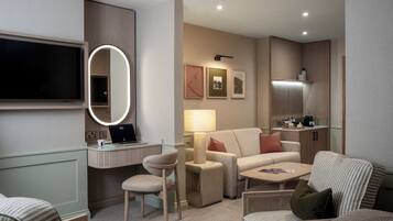 Quarto de hotel moderno com televisão de tela plana, escrivaninha com laptop, sofá confortável, mesa de centro pequena e área de cozinha compacta.