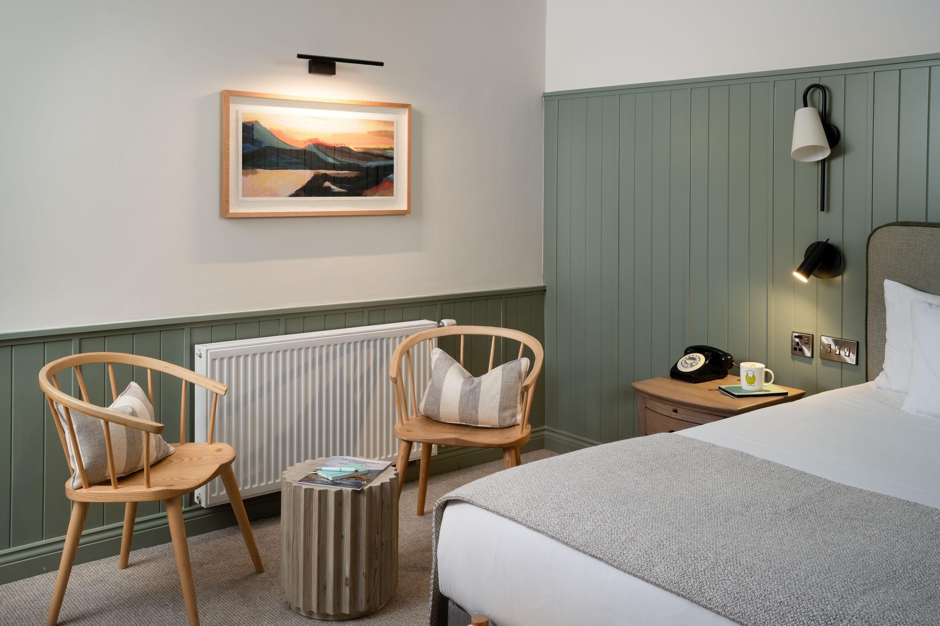 Quarto de hotel com uma cama, duas cadeiras de madeira, uma mesinha com abajur, um quadro na parede e um radiador.