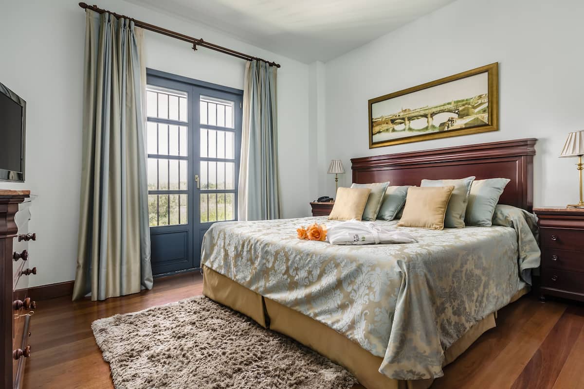 Um quarto com uma cama grande, uma cômoda de madeira, um quadro na parede e uma janela com cortinas.