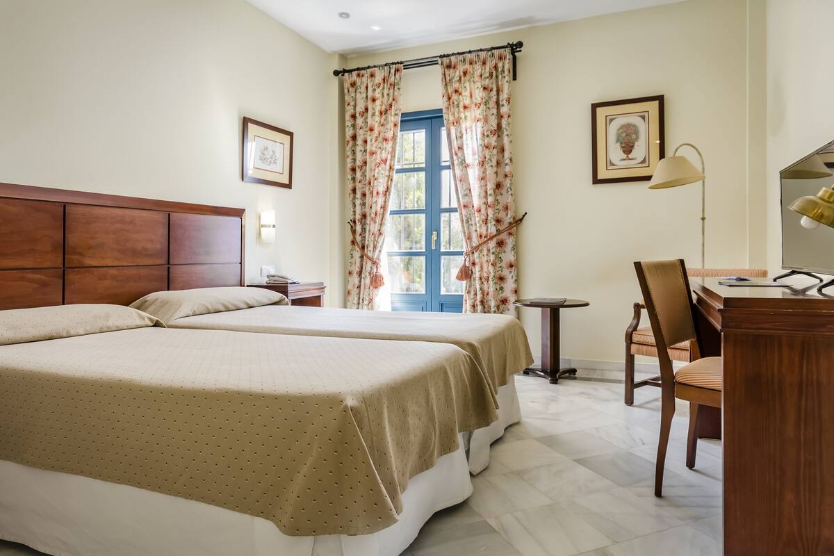 Quarto de hotel com duas camas, uma escrivaninha, uma cadeira, um abajur e uma janela com cortinas.