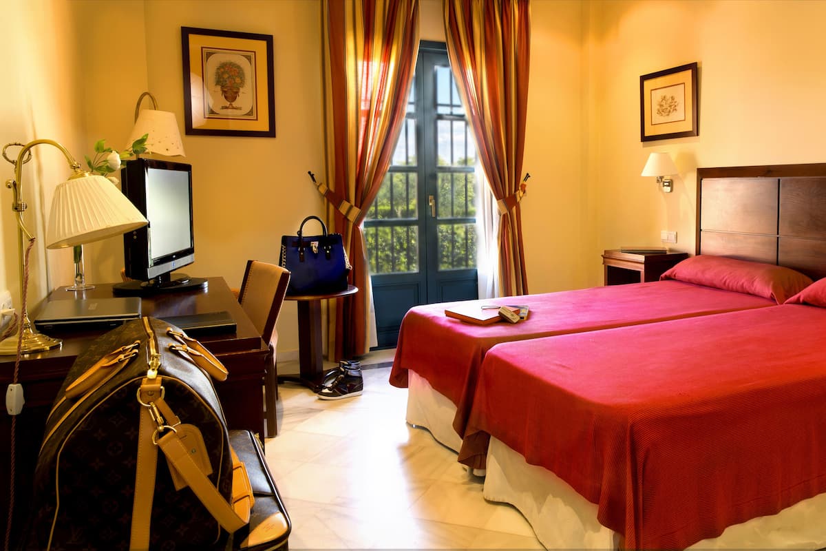 Quarto de hotel com cama de casal, escrivaninha com computador, cadeira, bolsa e janela com cortinas.