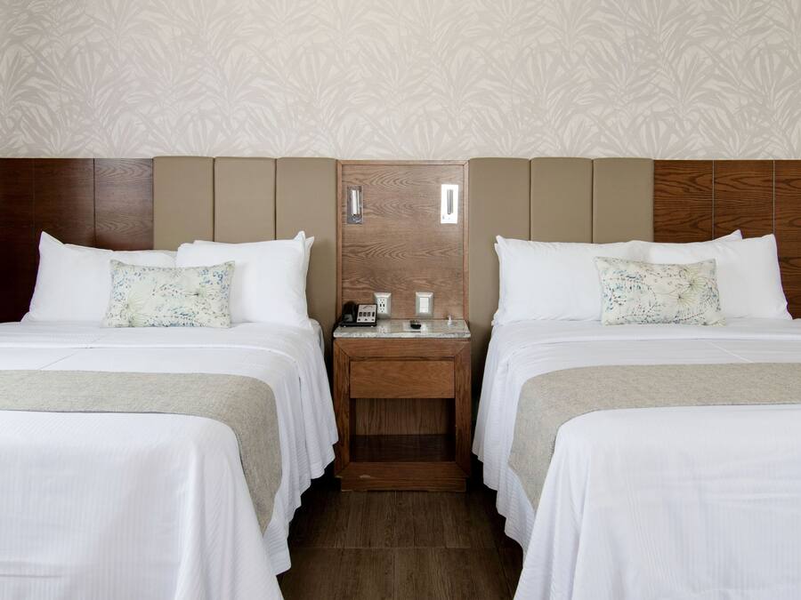 Quarto de hotel com duas camas, uma mesa de cabeceira de madeira com um telefone e uma pequena mesa, e um papel de parede estampado.