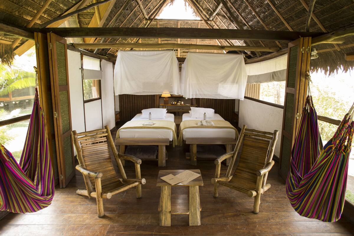 Quarto com duas camas, cadeiras de madeira, uma mesa pequena e um telhado de palha.