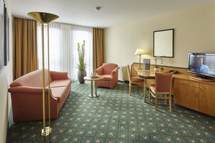 Quarto de hotel com carpete verde estampado, sofá, poltrona, escrivaninha com televisão e luminária.
