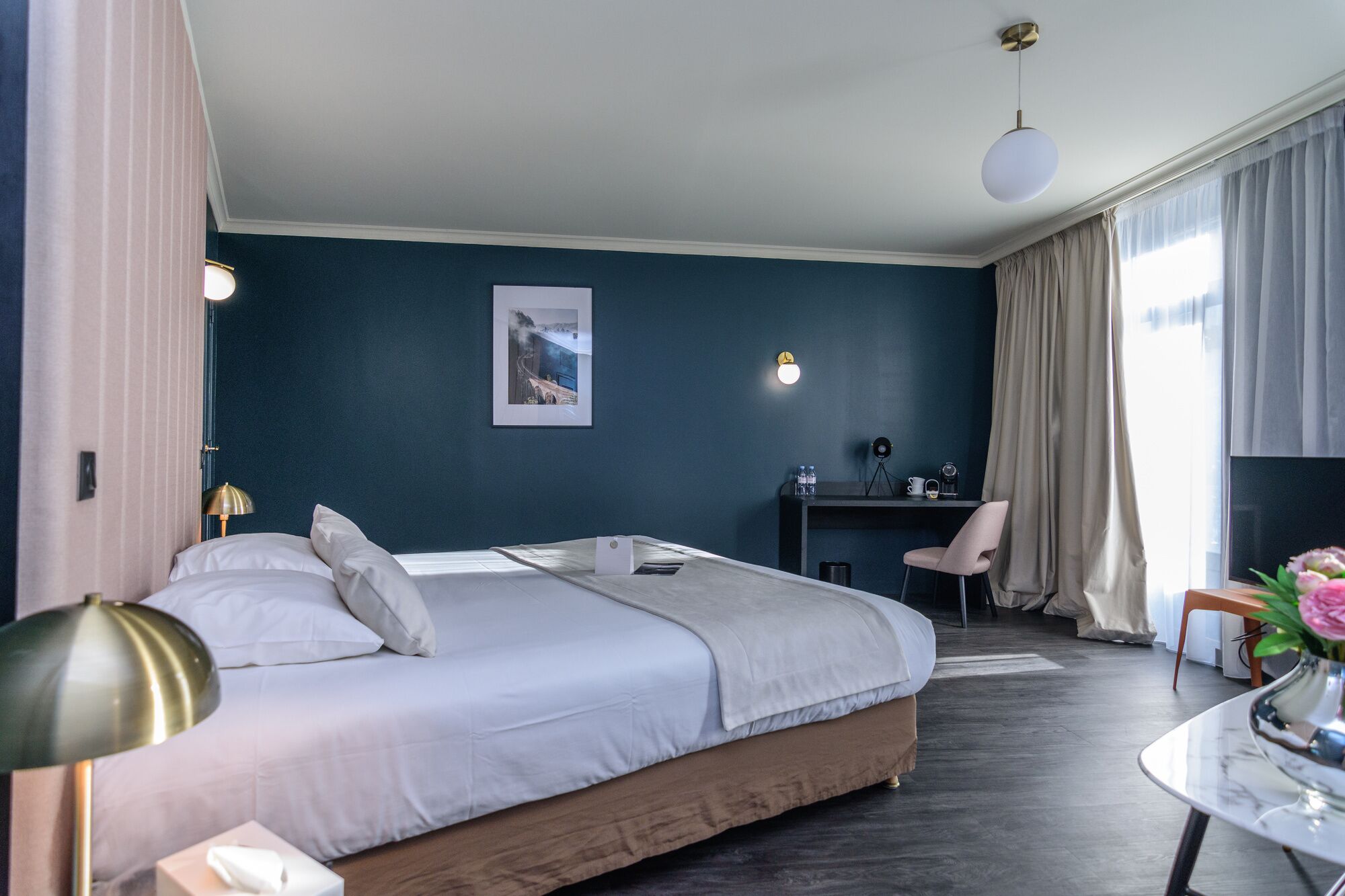 Quarto de hotel com uma cama grande, uma escrivaninha, uma cadeira e uma mesinha de centro com um vaso de flores.