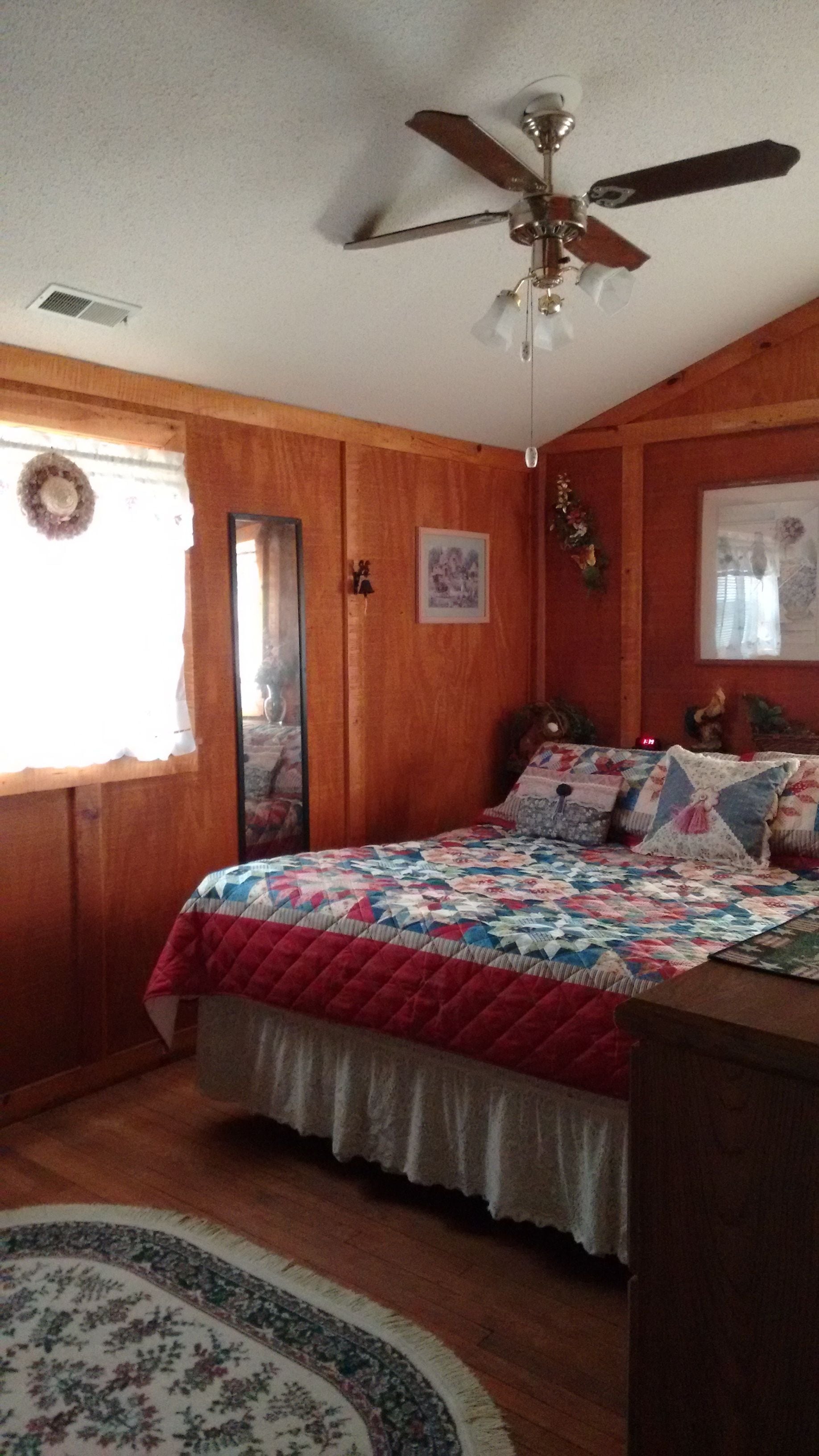 Quarto com piso de madeira, cama com colcha, ventilador de teto e janela com cortinas.