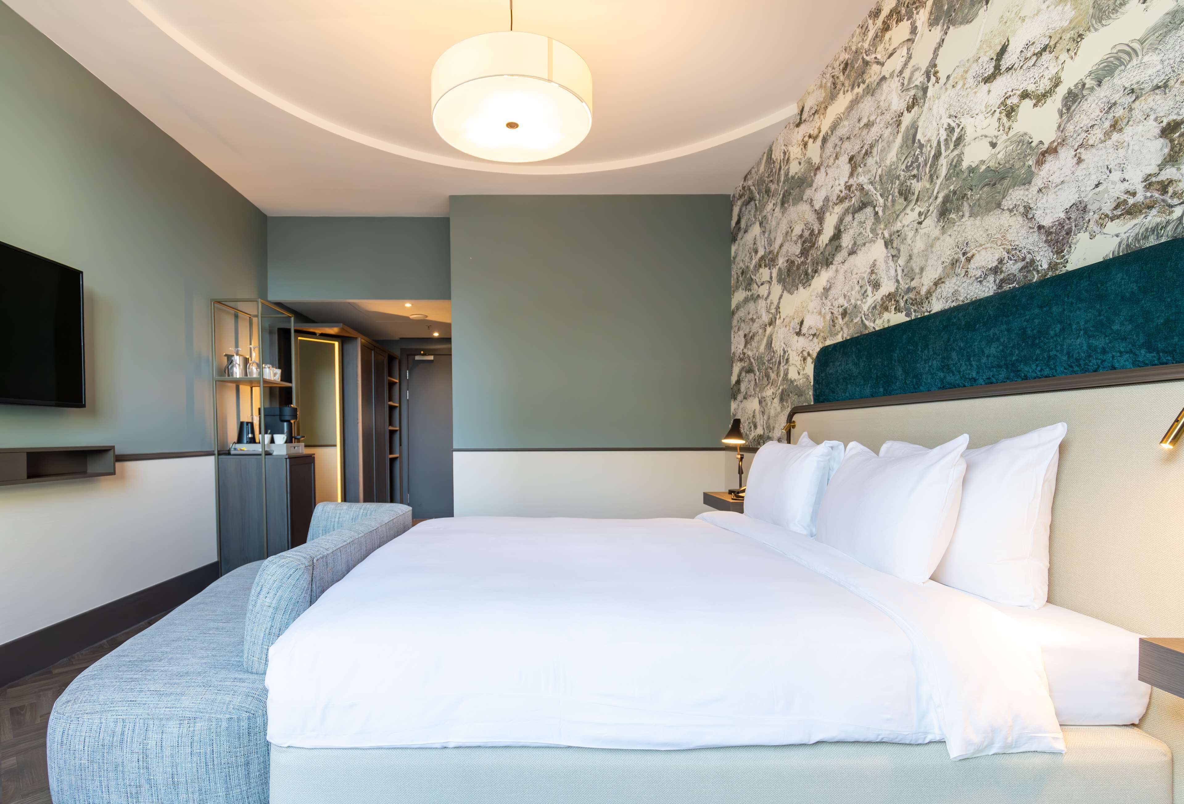 Quarto de hotel moderno com uma cama grande, uma televisão de tela plana na parede, uma mesinha de cabeceira com abajur e um guarda-roupa com espelho.