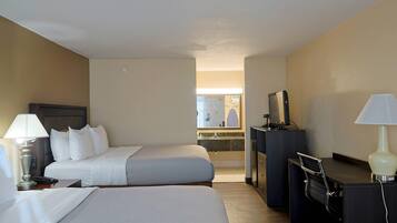 Quarto de hotel com duas camas, uma escrivaninha com computador, um espelho e um banheiro visível por uma porta aberta.
