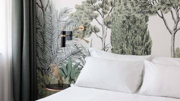 Quarto com cama, almofadas brancas, mesa de cabeceira com luminária e um mural de planta tropical na parede.