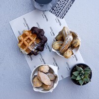 Waffles, churros e doces sobre uma mesa com um menu e uma pequena planta em vaso.