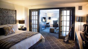 Quarto de hotel com cama, escrivaninha, cadeira, guitarra e televisão.