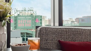 Vista da janela com um anúncio do Boston Red Sox ao fundo, uma mesa com uma tigela de cereais e um copo de cerveja, e um sofá com uma almofada vermelha.